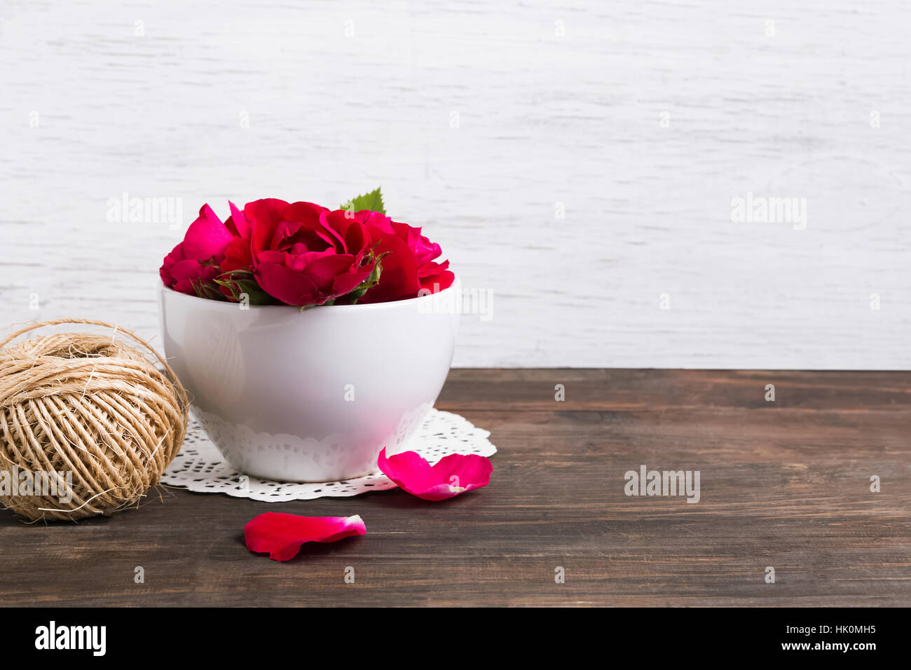 Little red roses in a cup, petals and jute rope on the wooden table ...