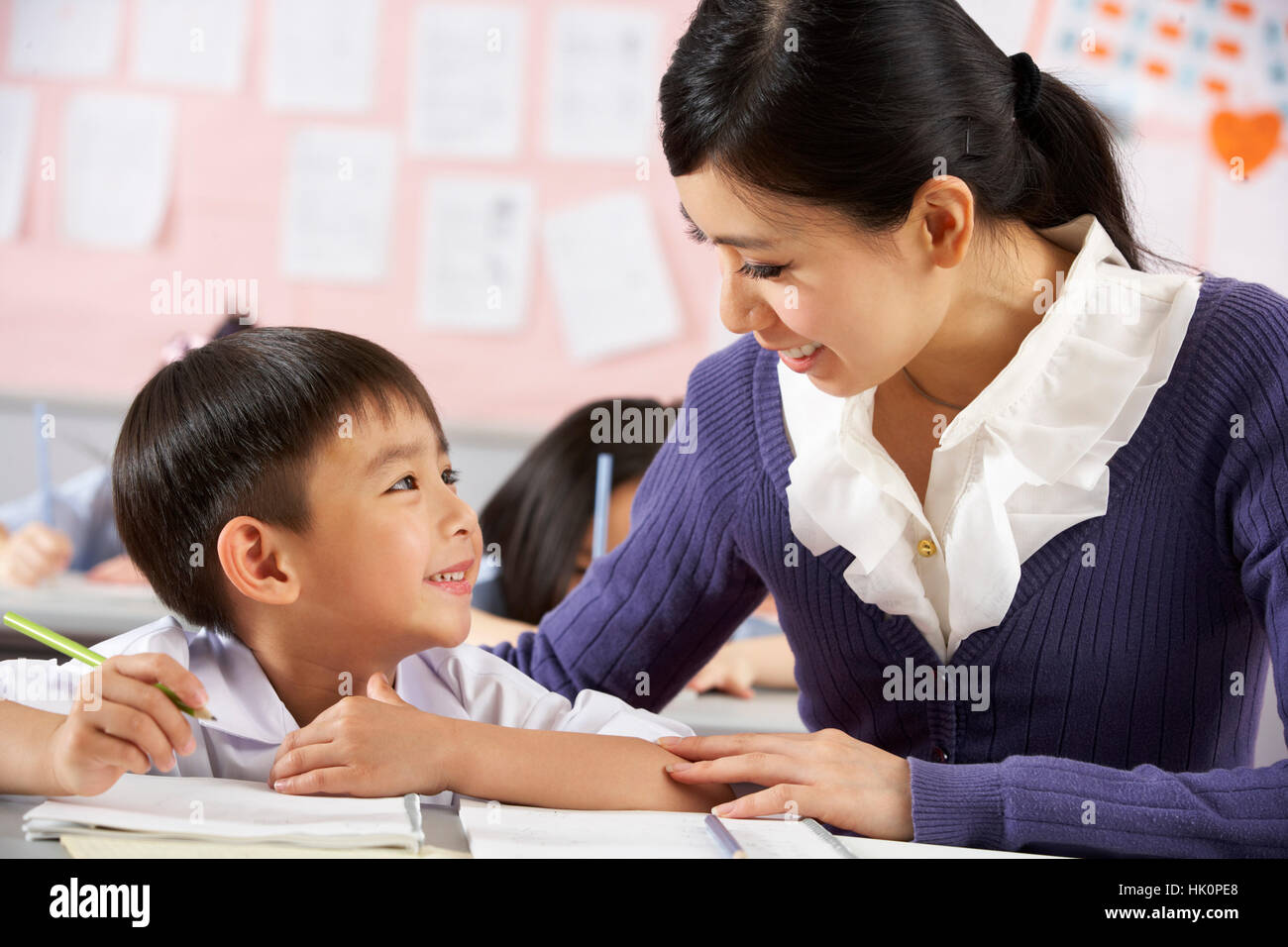 Teacher Helping Student Working At Desk In Chinese School Classroom ...