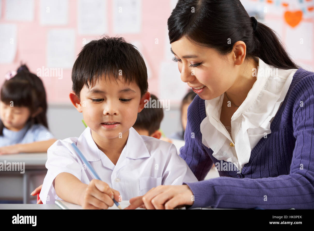 Teacher Helping Student Working At Desk In Chinese School Classroom ...