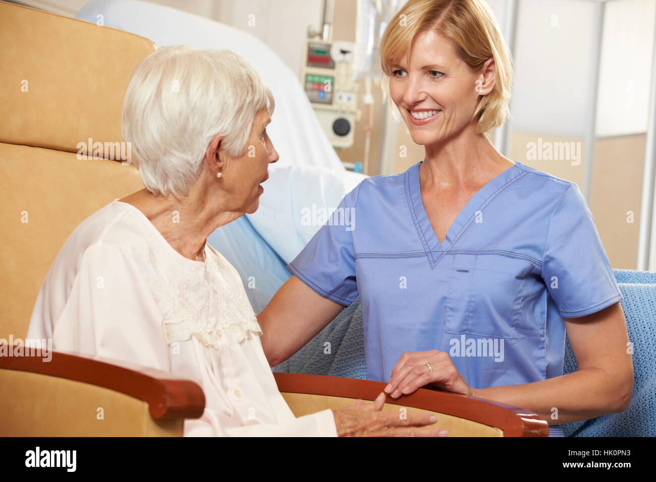 Nurse Taking To Senior Female Patient Seated In Chair By Hospital Bed ...