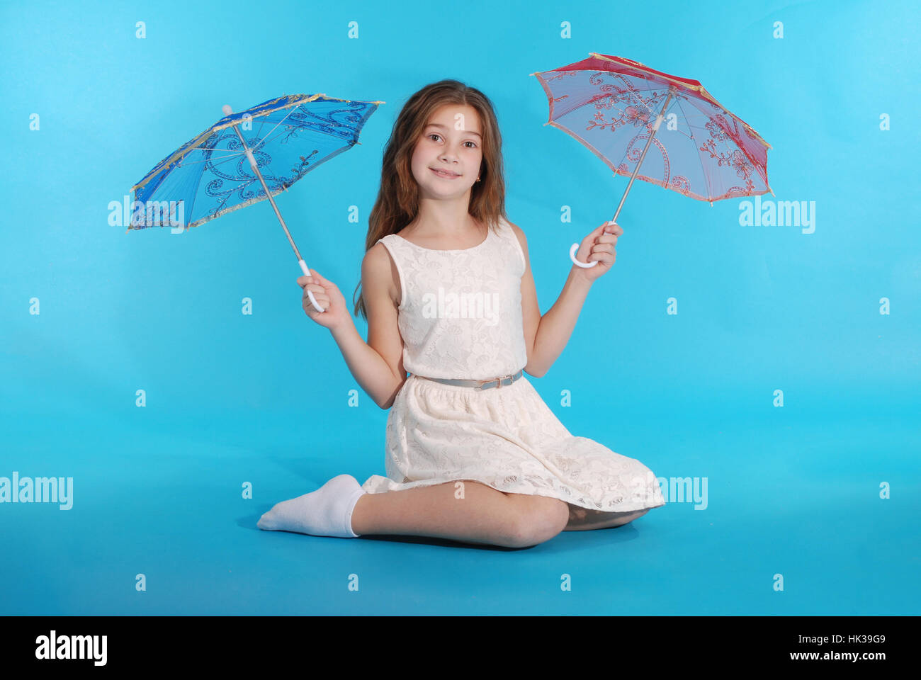 Cheerful Young Girl In White Dress With An Umbrella Sitting Against The Blue Background Stock