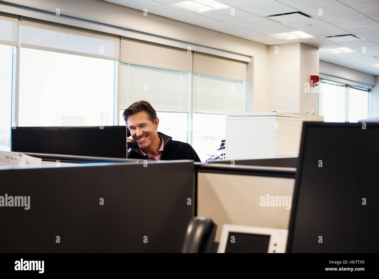 A man sitting in an office cubicle with a phone at his ear, smiling ...