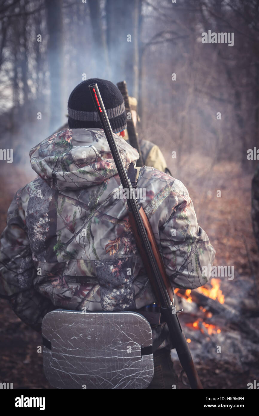 Hunter with gun at campfire in forest during hunting season. View from ...