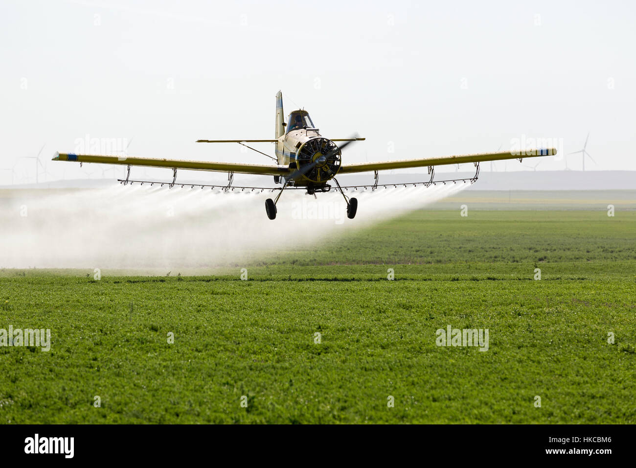 Crop duster flying and spraying over a green pea field with wind mills ...