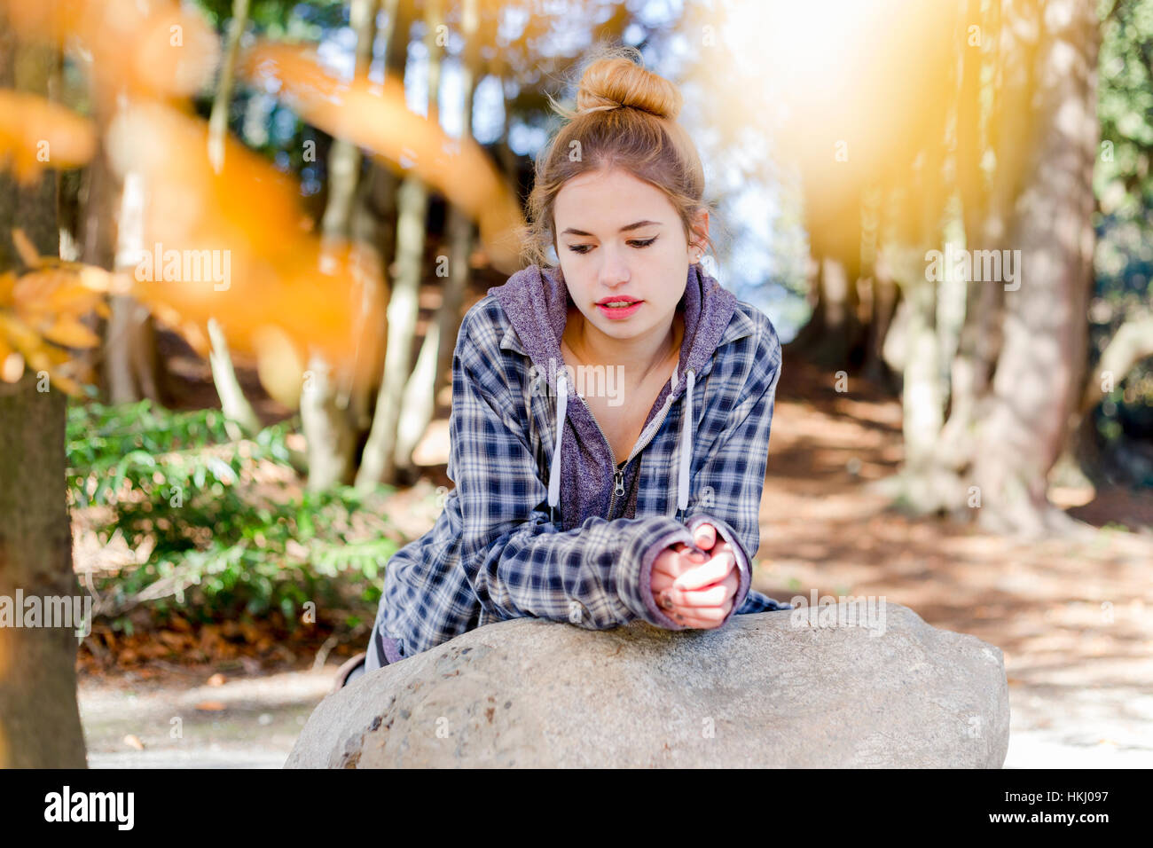 This young teenage girl hangs out alone in a park, sitting on a rock in ...