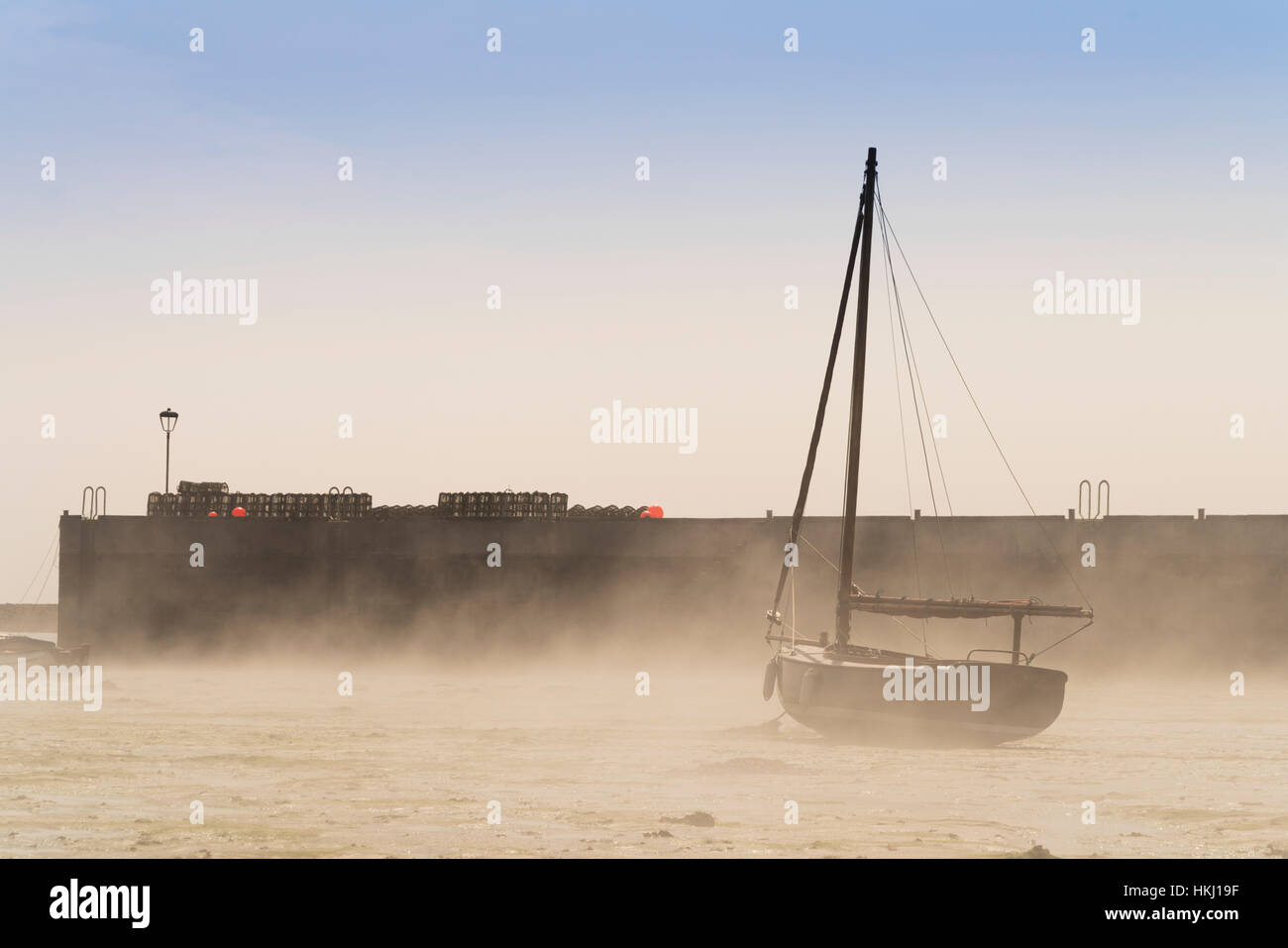Wind blows sand up on the beach against a sailboat sitting on the shore ...