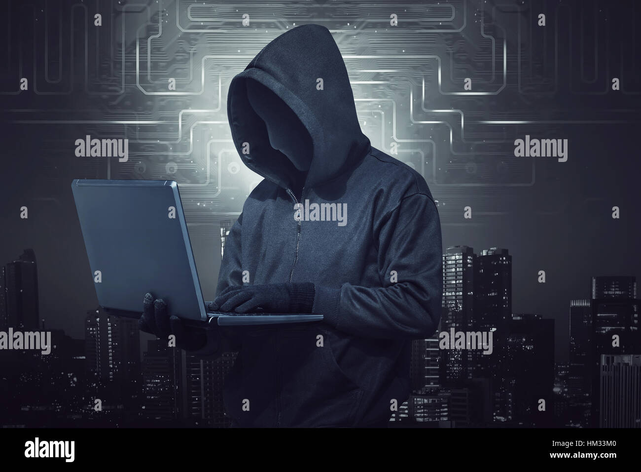 Hooded hacker holding laptop while typing against digital city ...