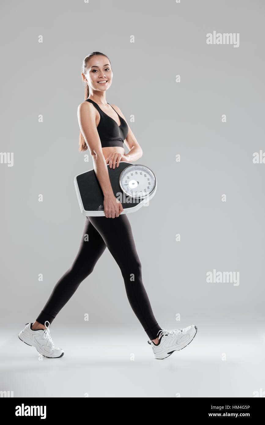 Happy cute young woman athlete walking and holding weighing scale over ...
