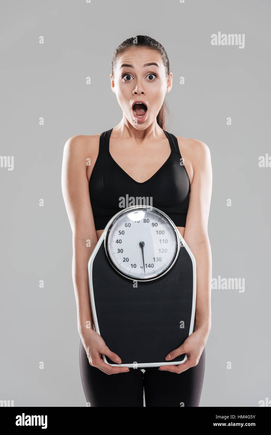 Shocked astonished young woman athlete holding weighing scale over gray ...