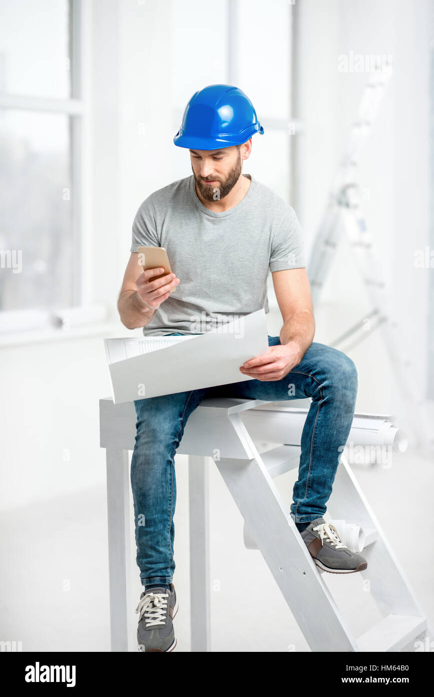 Handsome repairman or builder in helmet working with drawings and phone ...