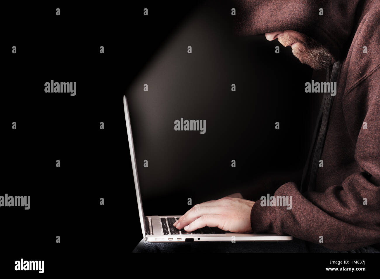 Male computer hacker wearing a hooded top leaning over a laptop in the ...