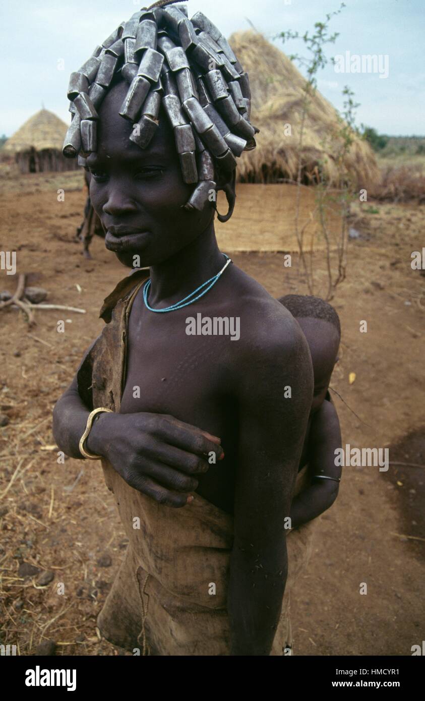 Young Mursi woman with a traditional hairstyle and her baby swaddled on ...