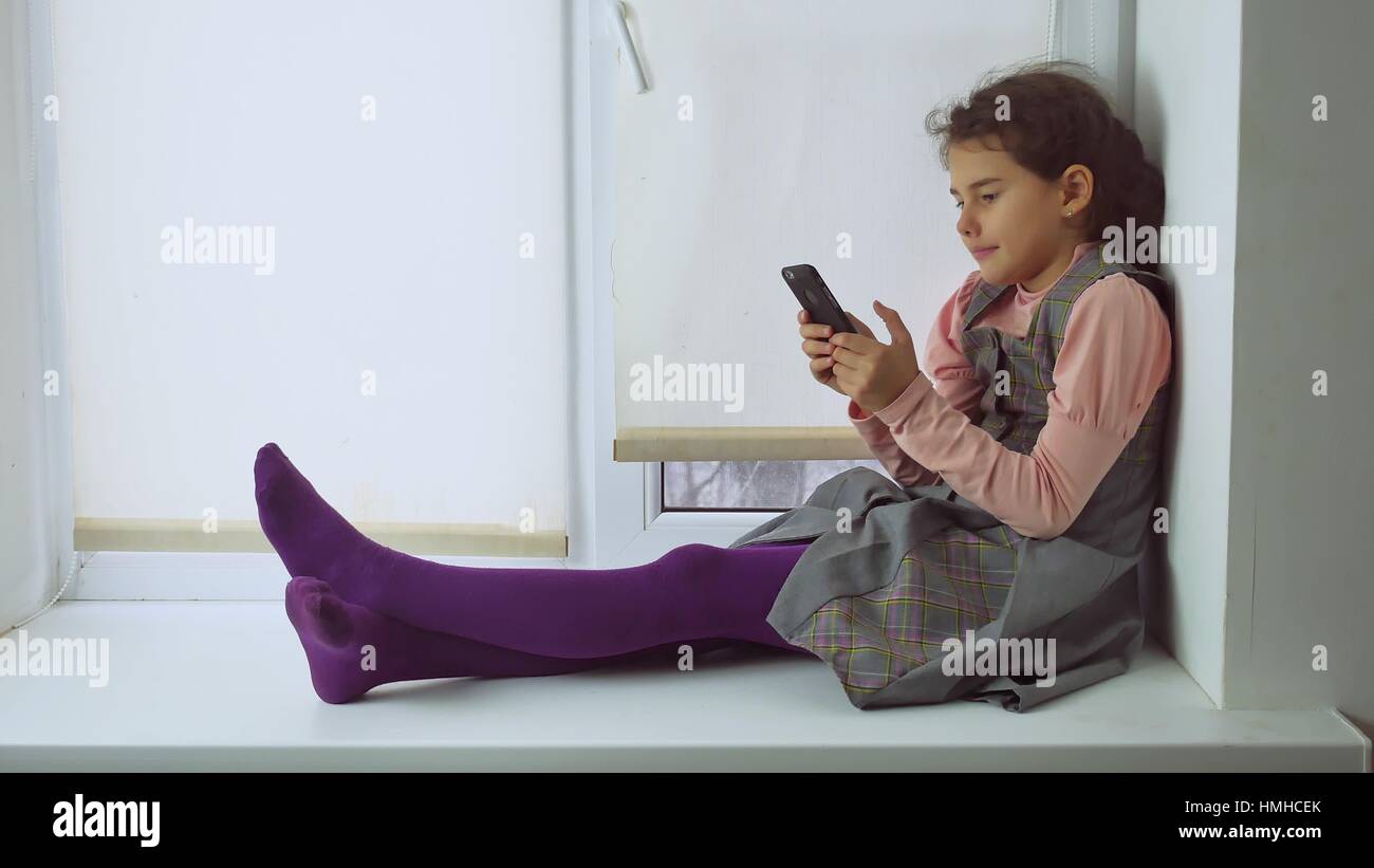 teen girl sitting on a window sill plays the online game for smartphone ...