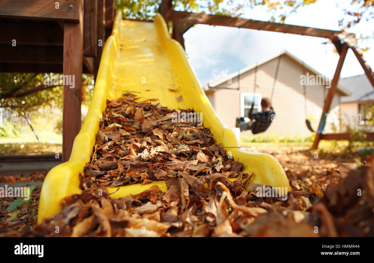 A play-set with fall leaves on the slide. A boy is swinging on the ...