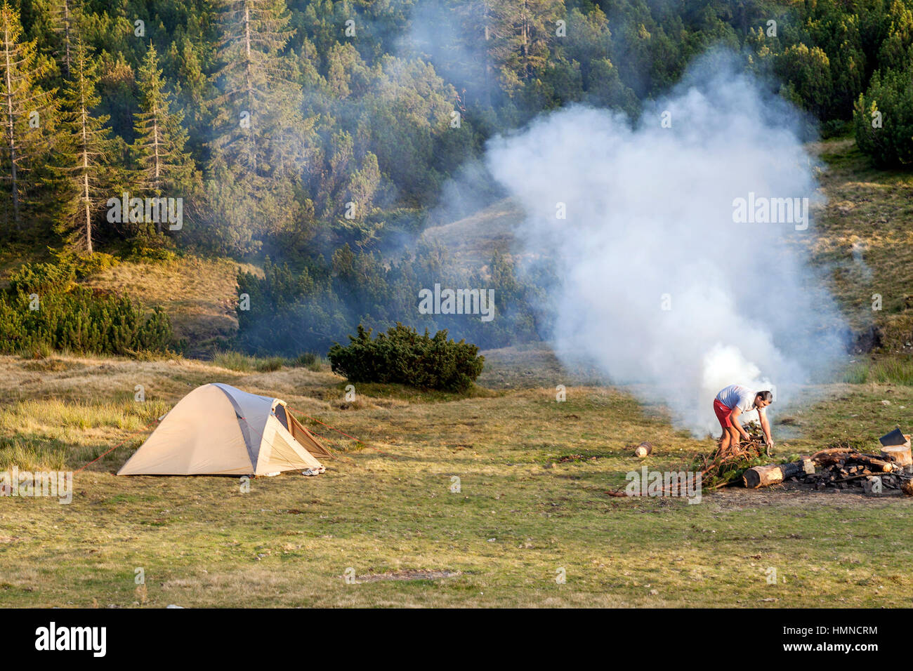 View with a tent, camping fire wit smoke and tourist hiker in summer ...