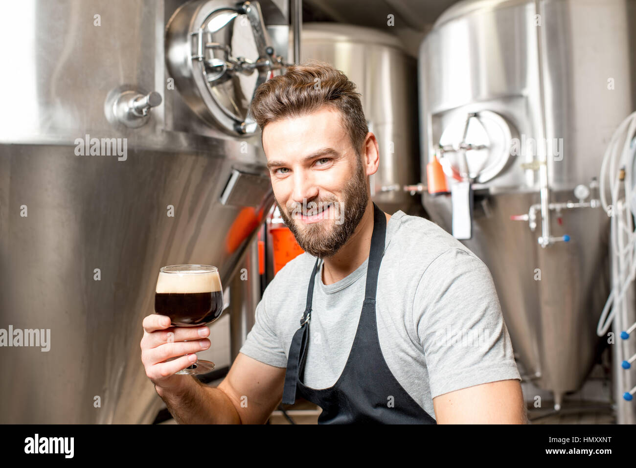 Handsome brewer in uniform tasting beer at the brewery with metal ...