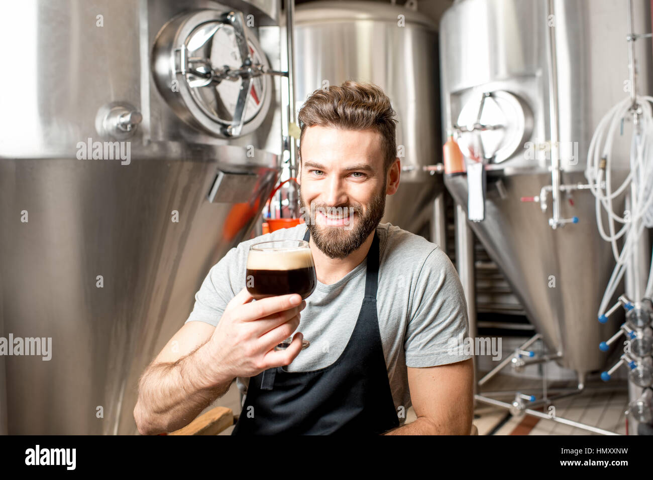 Handsome brewer in uniform tasting beer at the brewery with metal ...