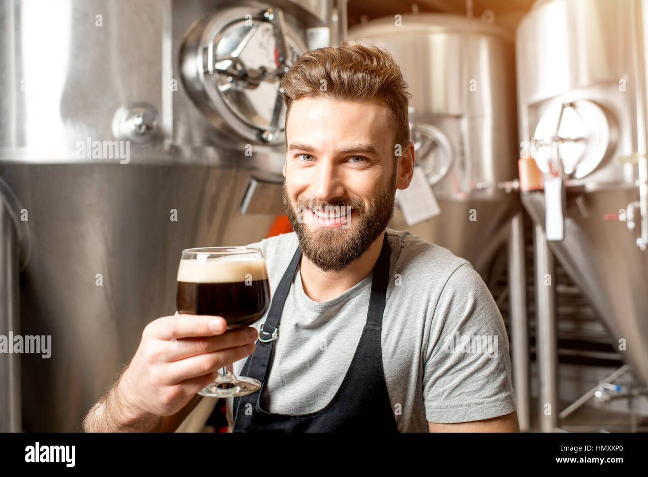 Handsome brewer in uniform tasting beer at the brewery with metal ...