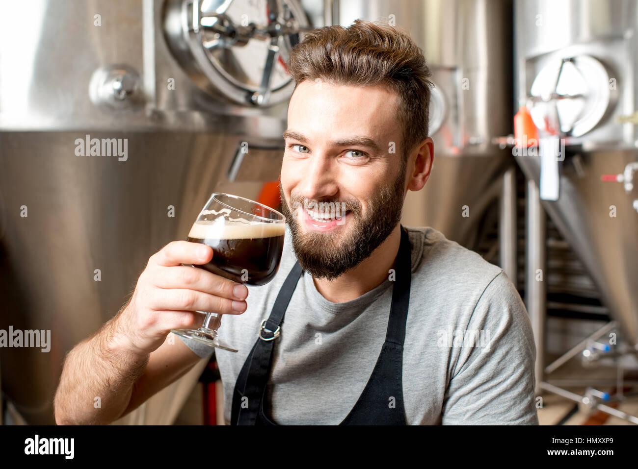 Handsome brewer in uniform tasting beer at the brewery with metal ...