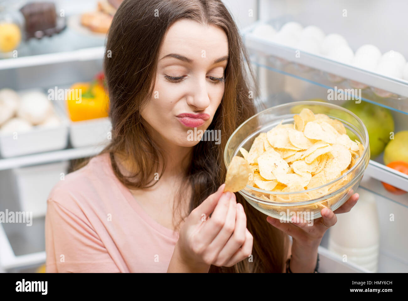 Young woman eating potato chips near the refrigerator full of healthy ...