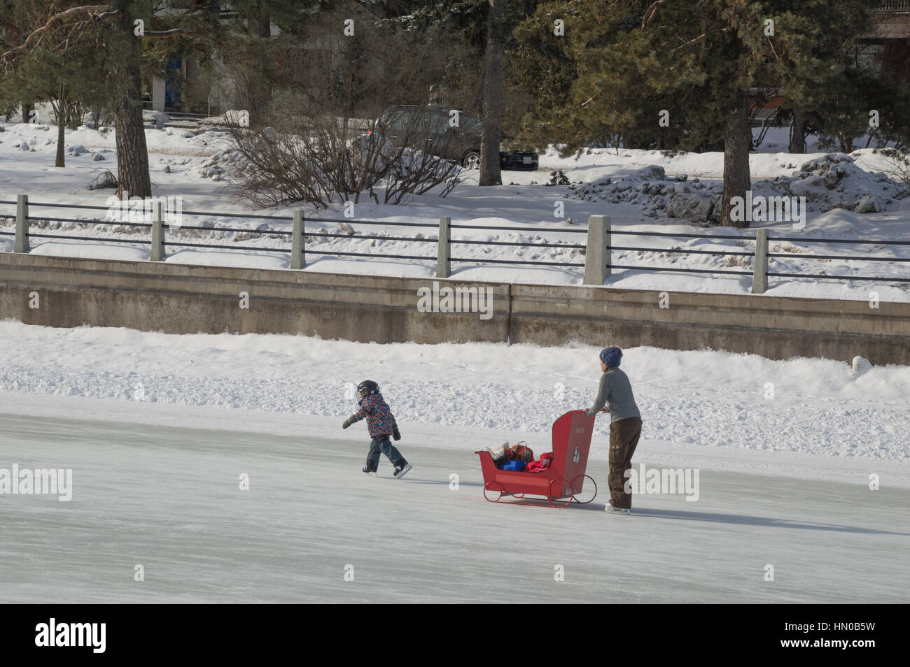 Mom pushing a red sleigh stroller, with young child skating on Rideau ...