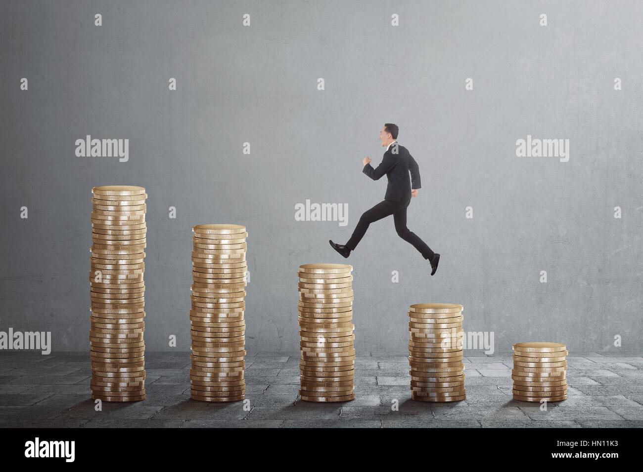Happy asian businessman jump on between coin stacks against grey ...