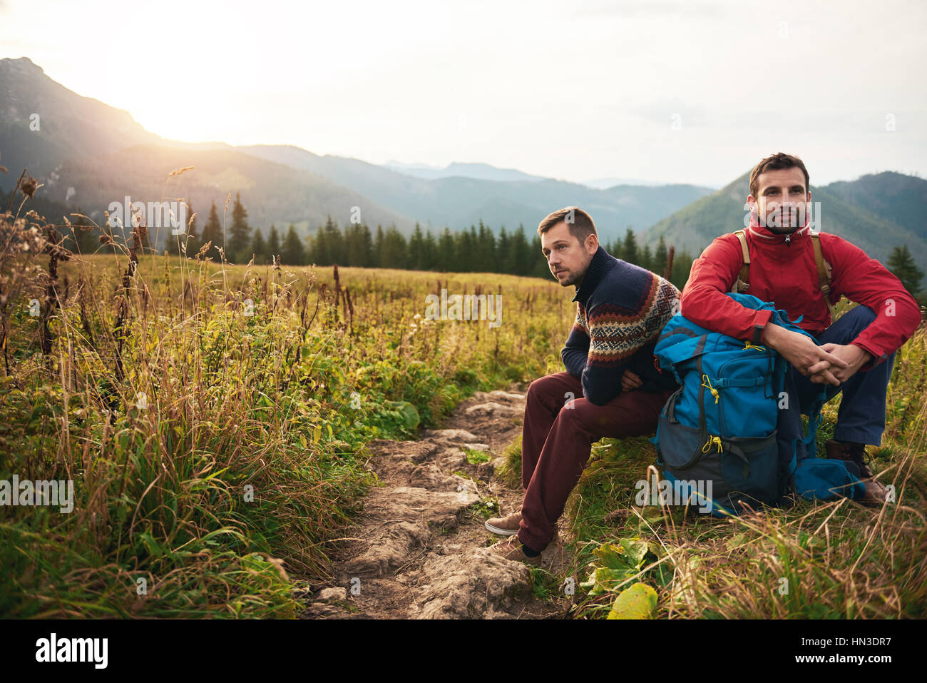 Two young men in hiking gear sitting on a path at sunrise while taking ...