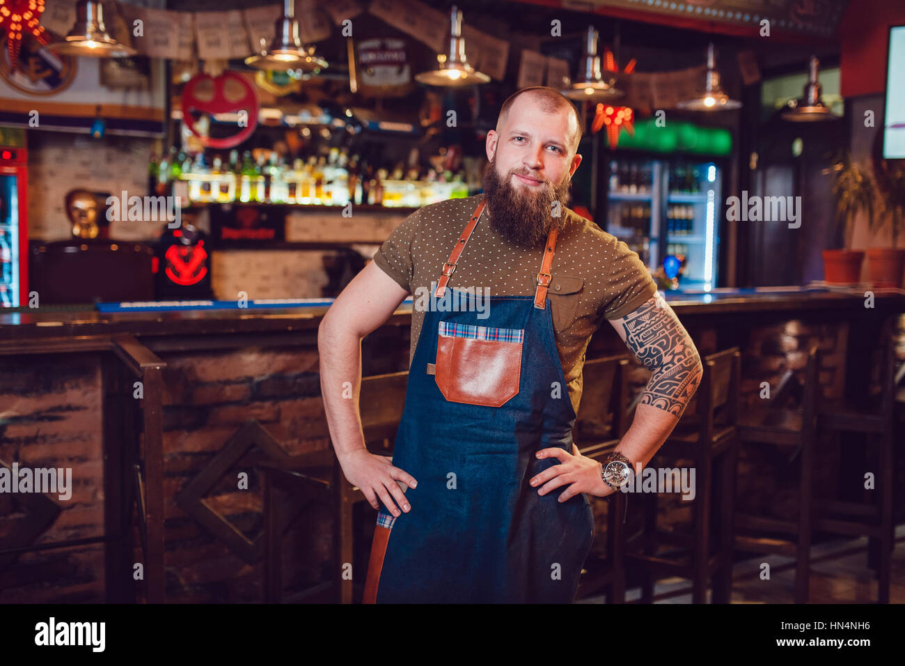 Bearded barman with tattoos and watches wearing an apron standing near ...