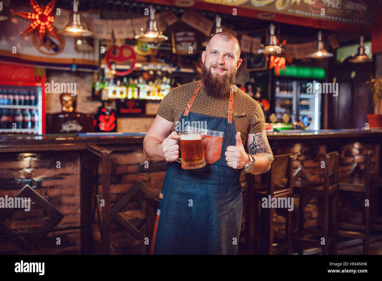 Bearded barman with tattoos wearing an apron standing near the bar and ...