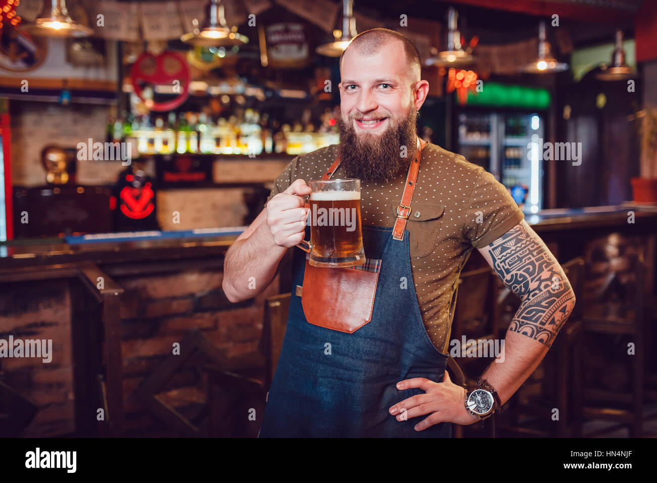 Bearded barman with tattoos wearing an apron standing near the bar and ...