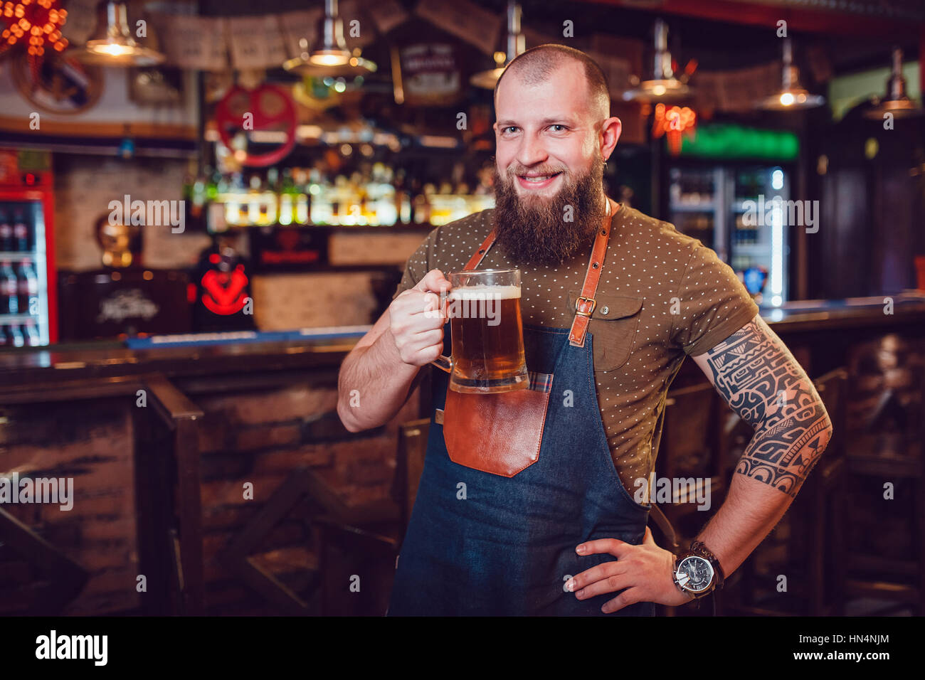 Bearded barman with tattoos wearing an apron standing near the bar and ...
