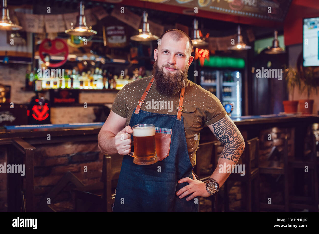 Bearded barman with tattoos wearing an apron standing near the bar and ...