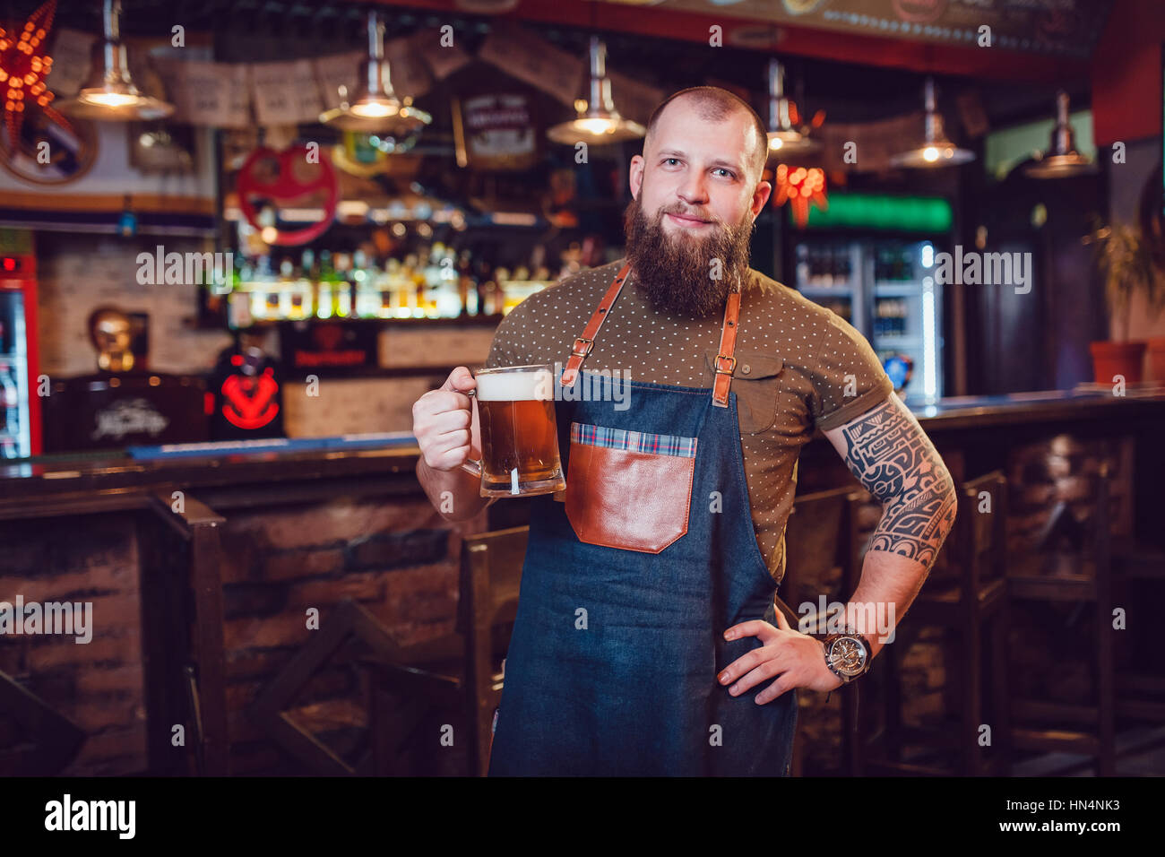 Bearded barman with tattoos wearing an apron standing near the bar and ...
