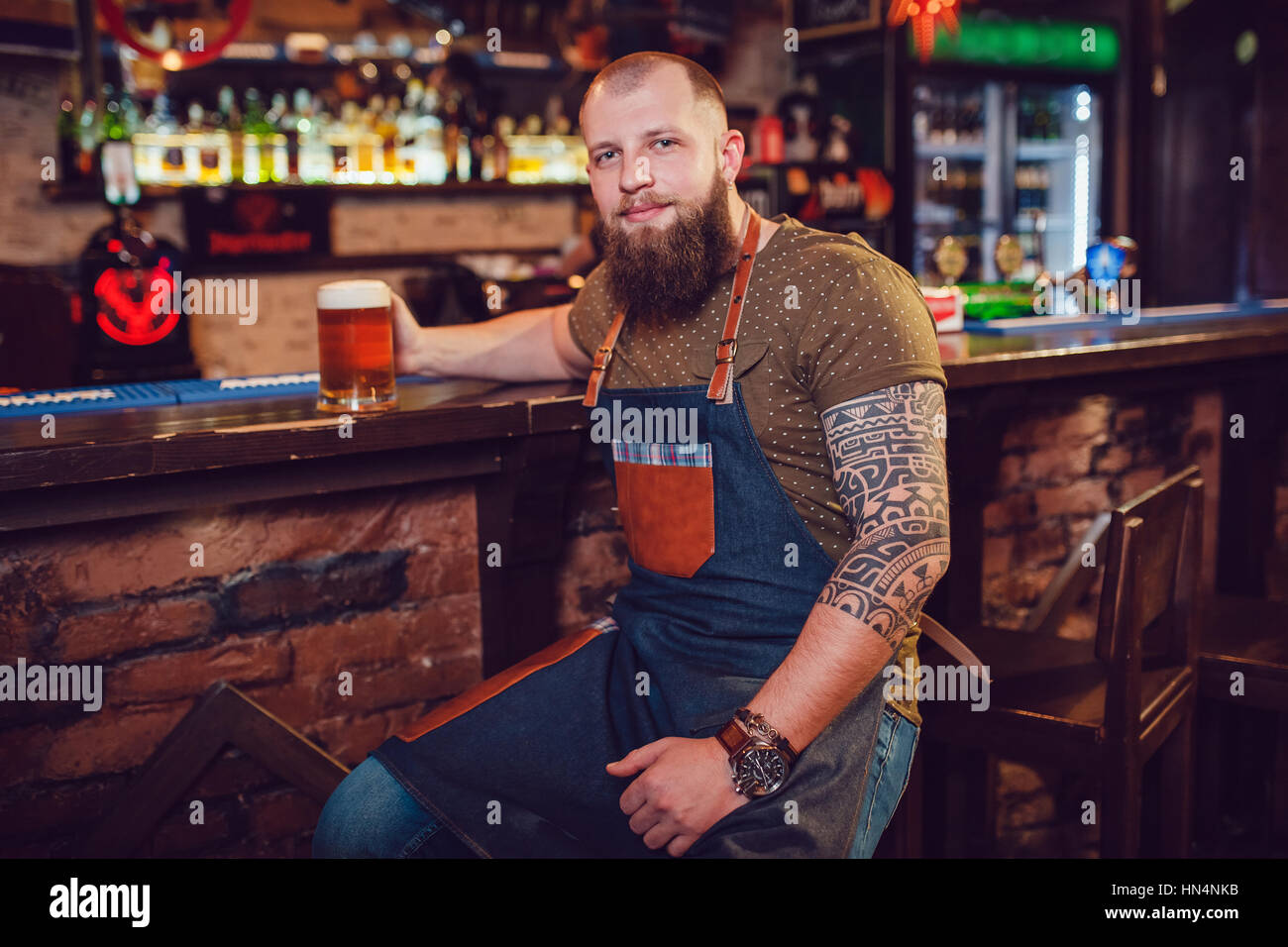 Bearded barman with tattoos wearing an apron sitting near the bar and ...