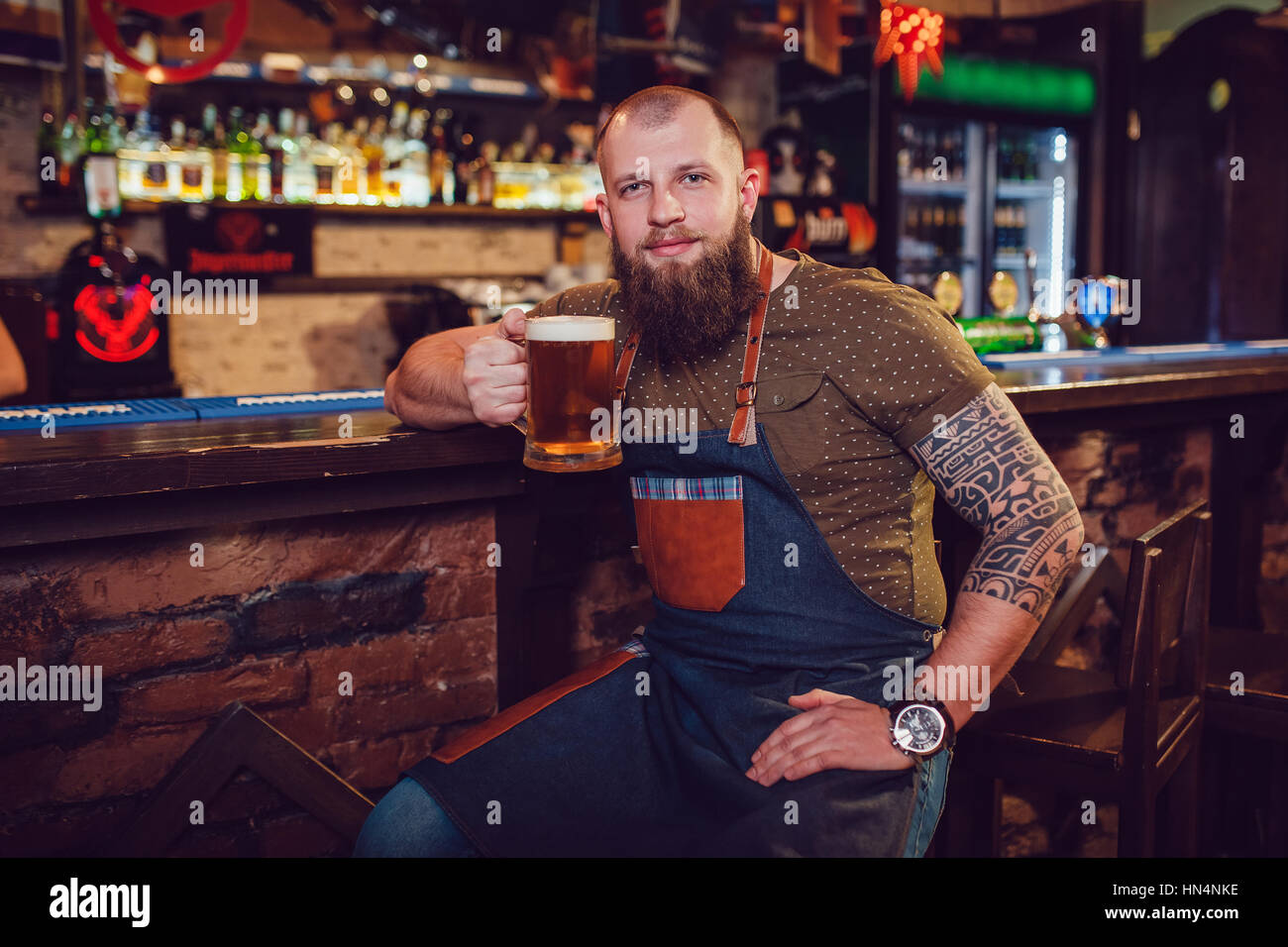 Bearded barman with tattoos wearing an apron sitting near the bar and ...
