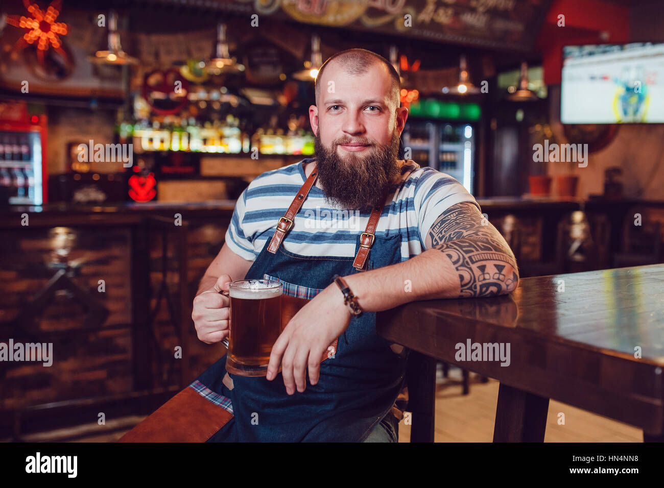 Bearded barman with tattoos wearing an apron sitting in bar and holding ...
