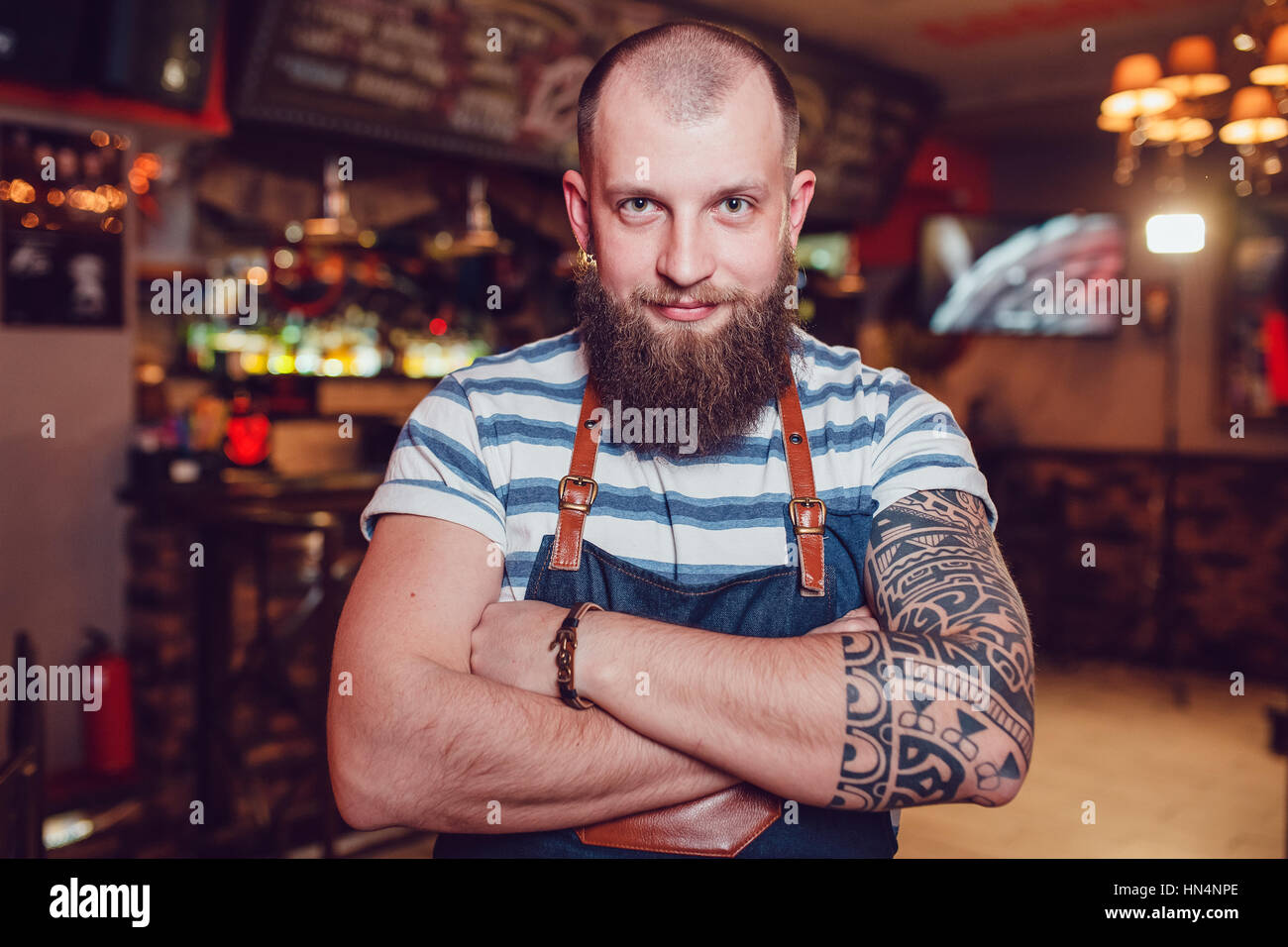 Bearded barman with tattoos wearing an apron standing at the bar with ...