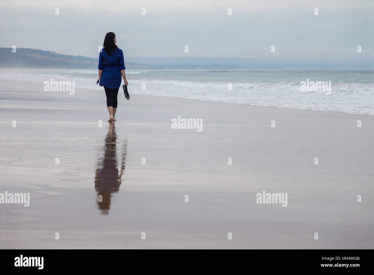 Young woman walking alone in a deserted beach reflected on the wet sand ...