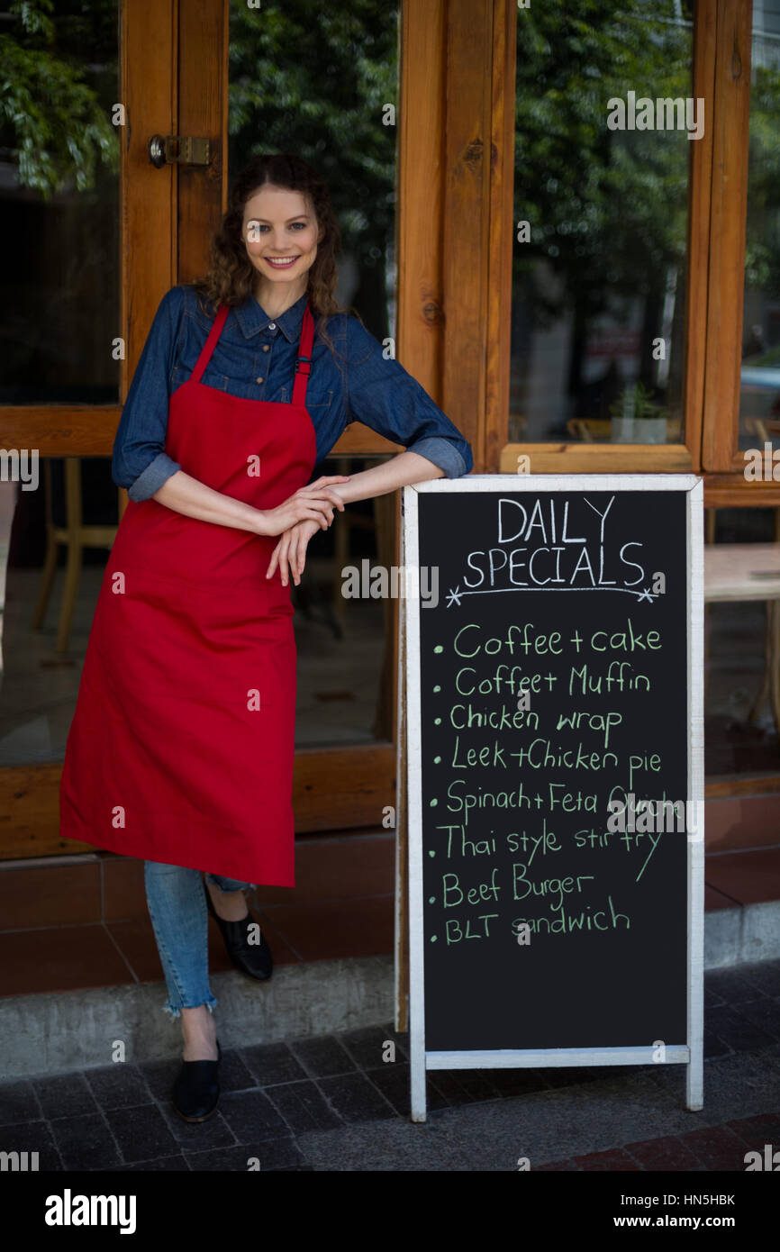Portrait of smiling waitress leaning on menu board outside the cafe ...