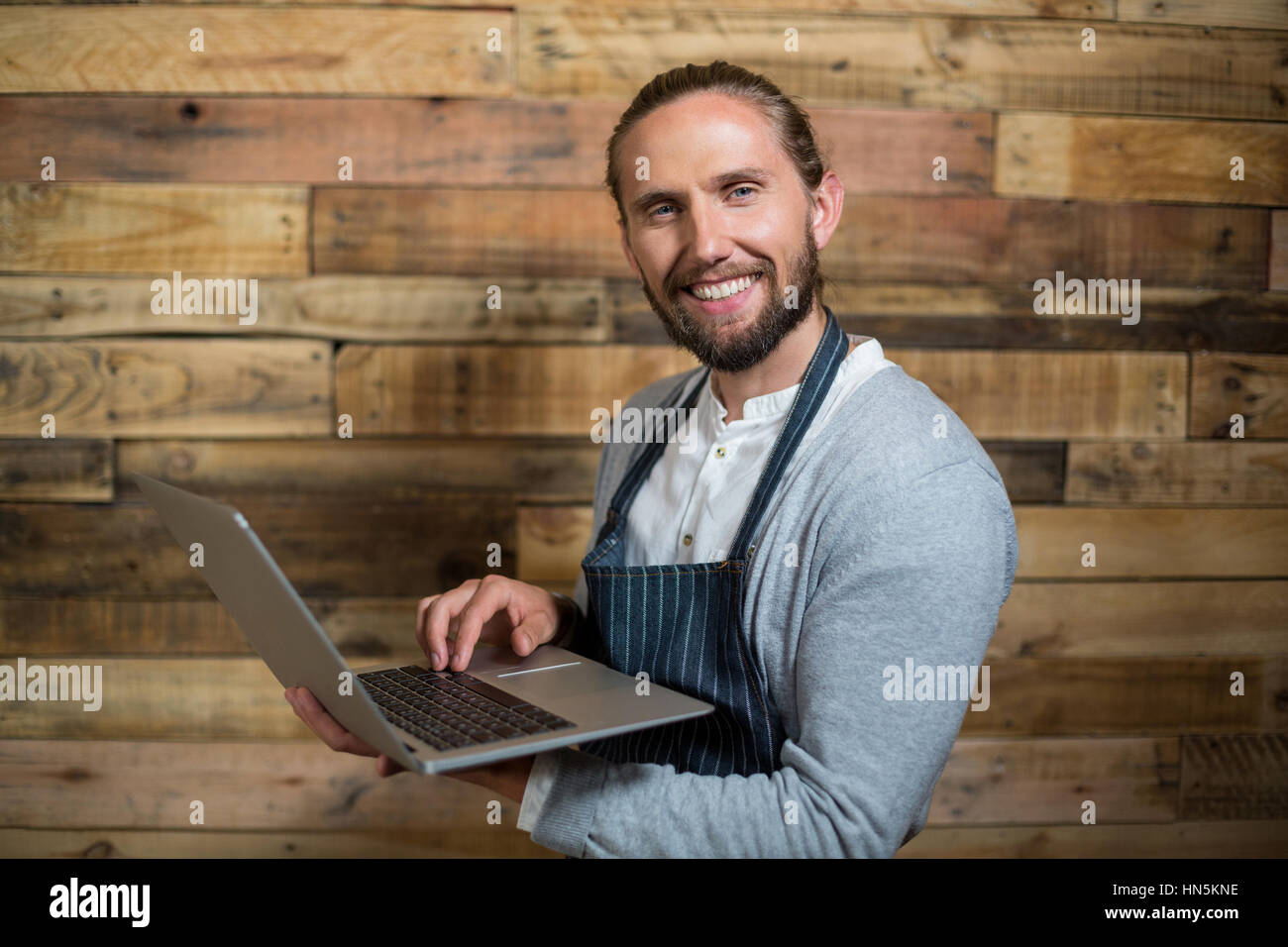 Portrait of smiling waiter using laptop against wooden wall in cafÃƒÂ ...