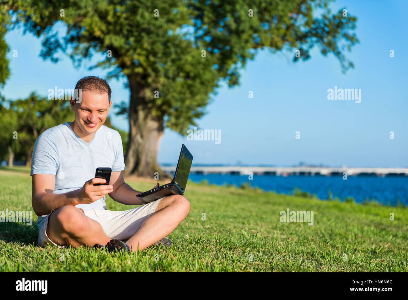 Young man reading smartphone and laughing sitting outside in park with ...
