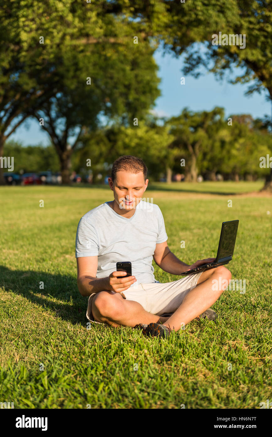 Young man looking at phone and smiling sitting outside in green grass ...