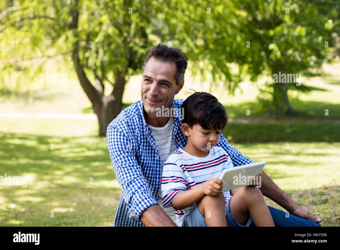 Boy sitting on his fathers lap and using digital tablet in park on a ...