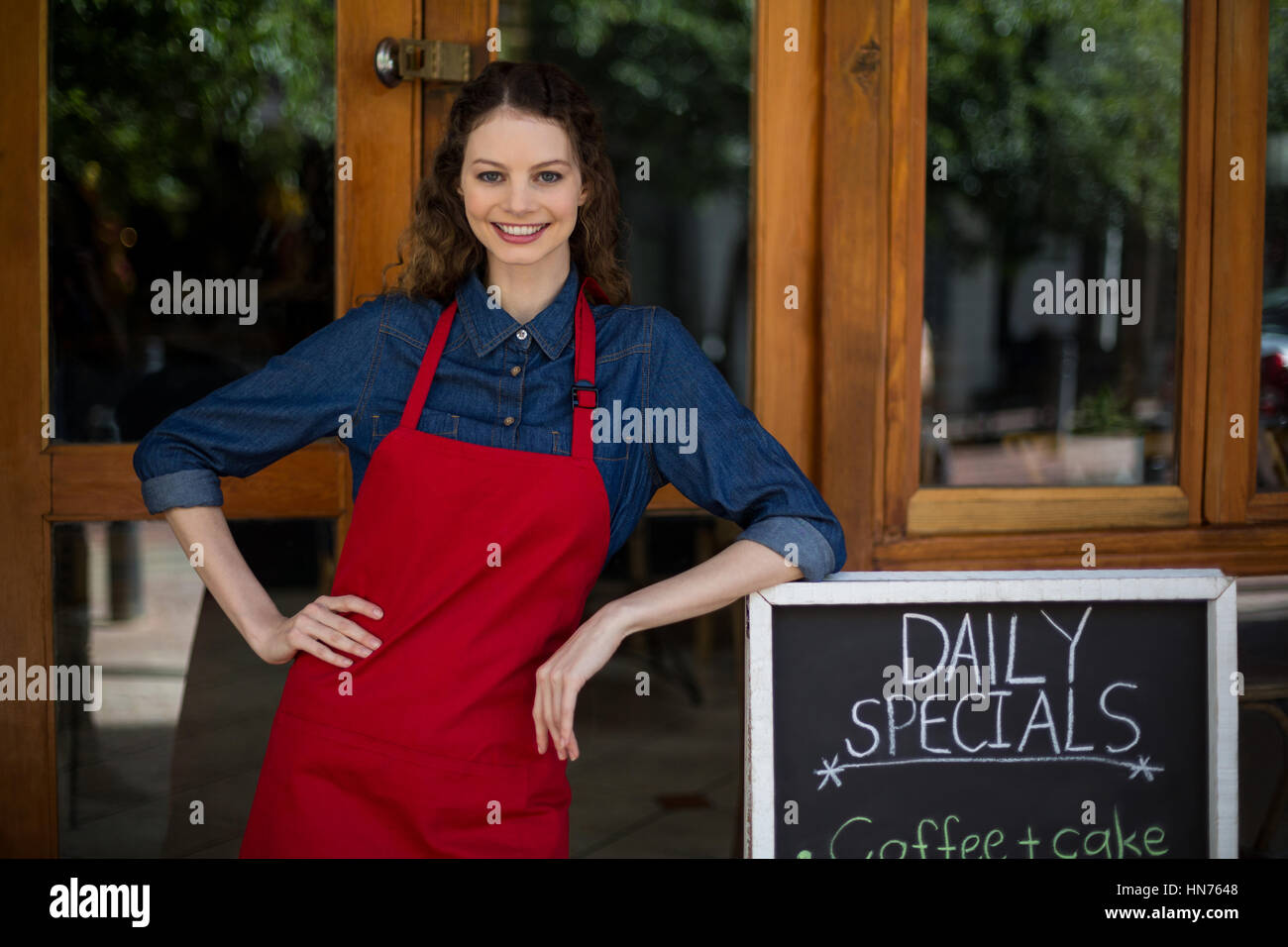 Portrait of smiling waitress leaning on menu board outside the cafe ...