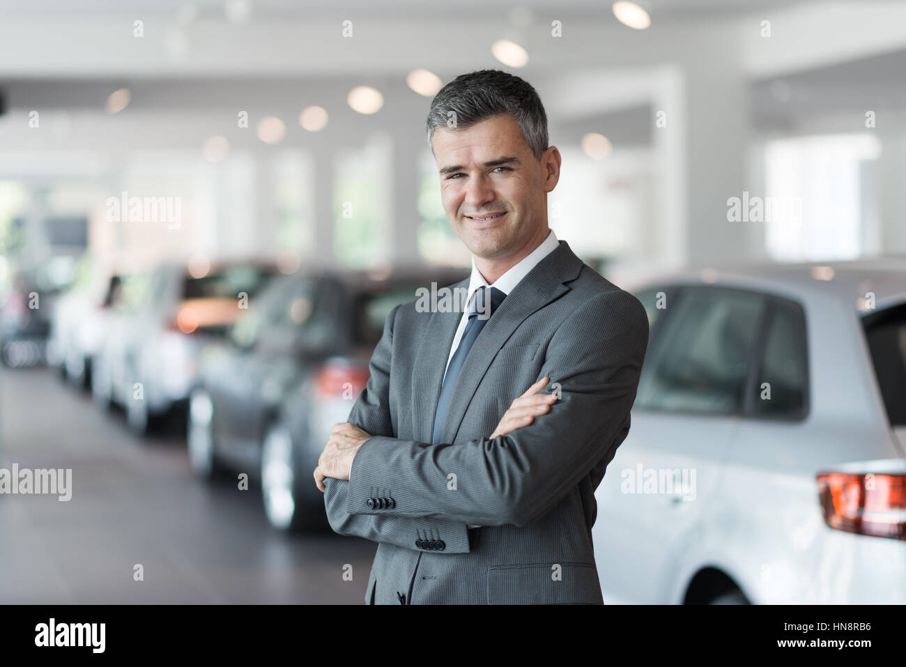 Confident smiling car salesman at the showroom, he is standing with ...