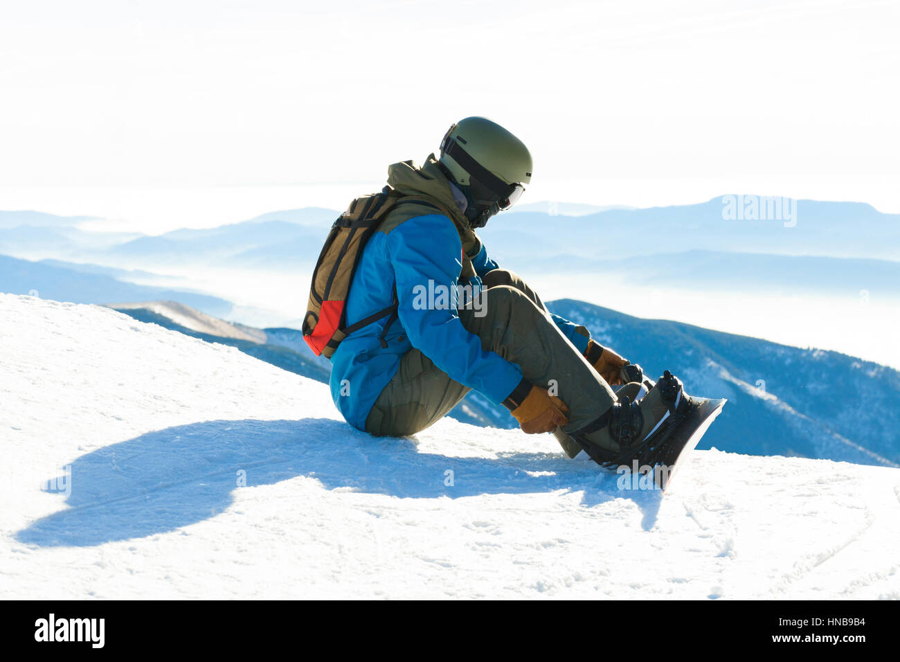 Snowboarder in helmet sitting at the top of a mountain and tightening ...