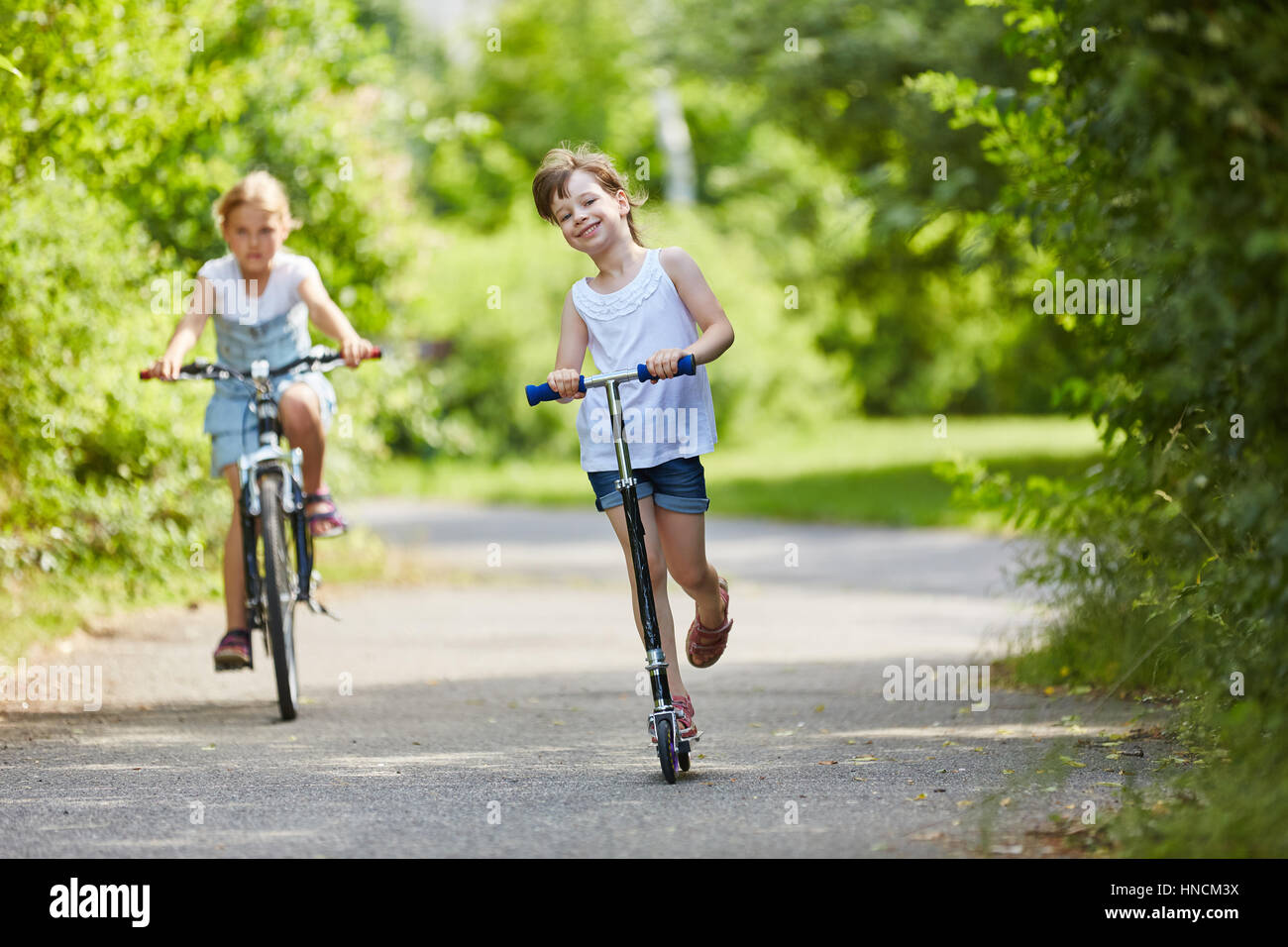 Girls drive scooter and ride bike in the park during summer vacation ...