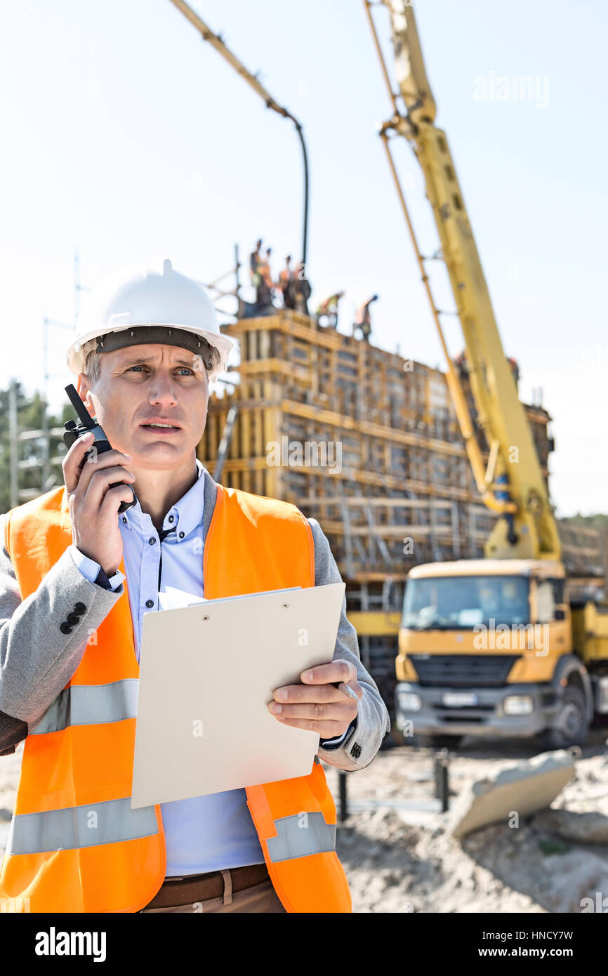 Male supervisor using walkie-talkie while holding clipboard at ...