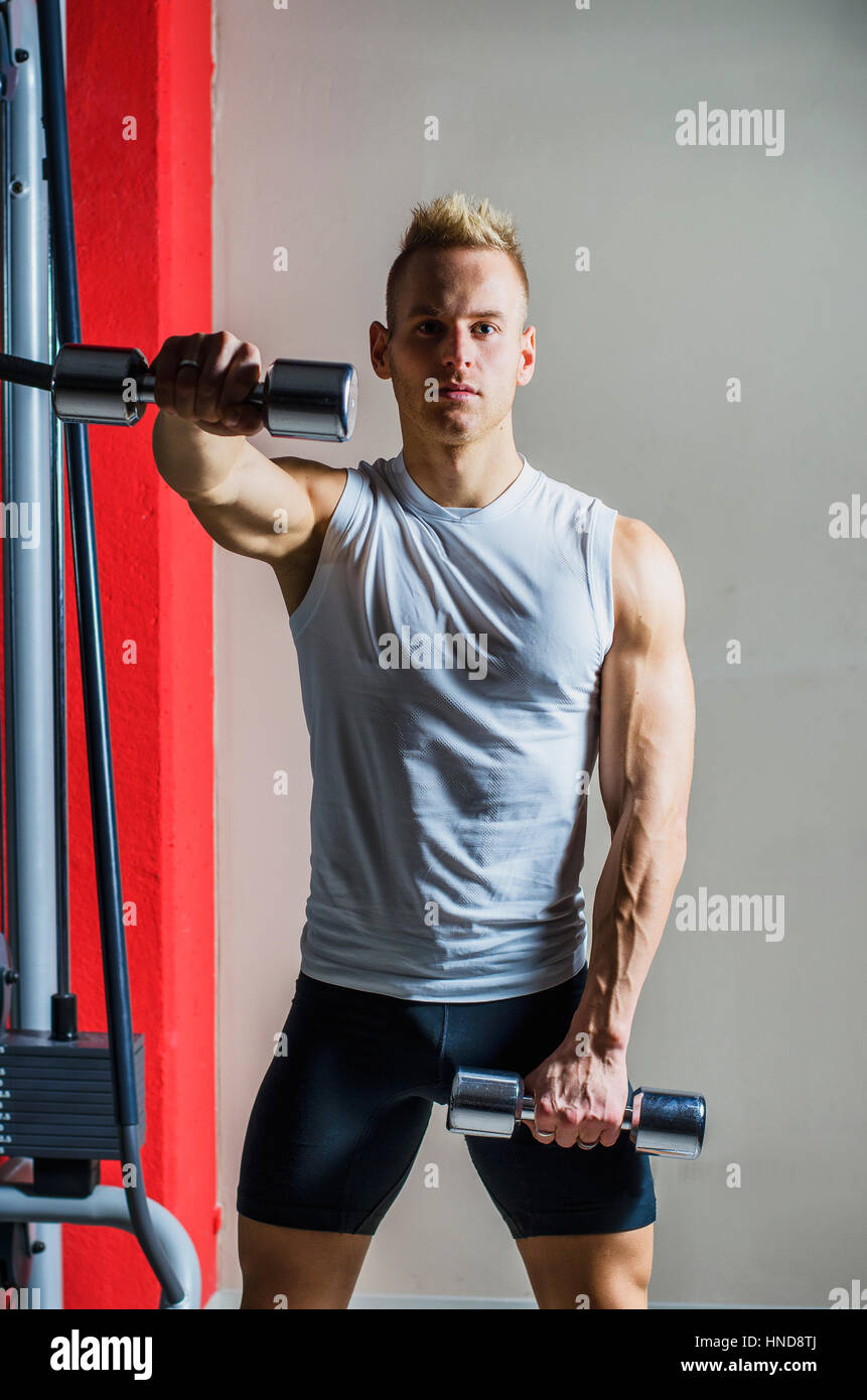 Muscular blond young man standing in gym working out with dumbbells ...