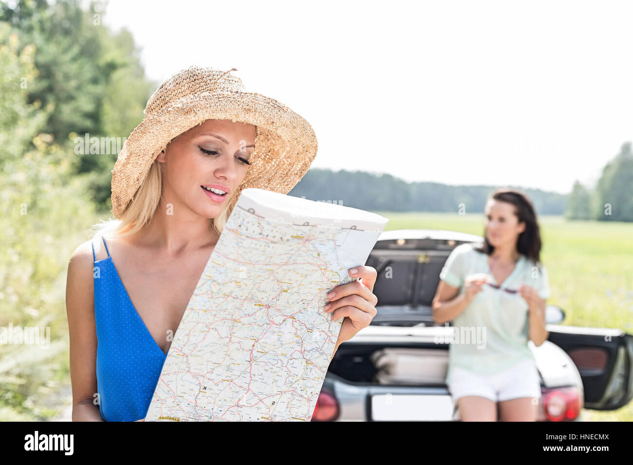Woman reading map while friend leaning on convertible in background ...